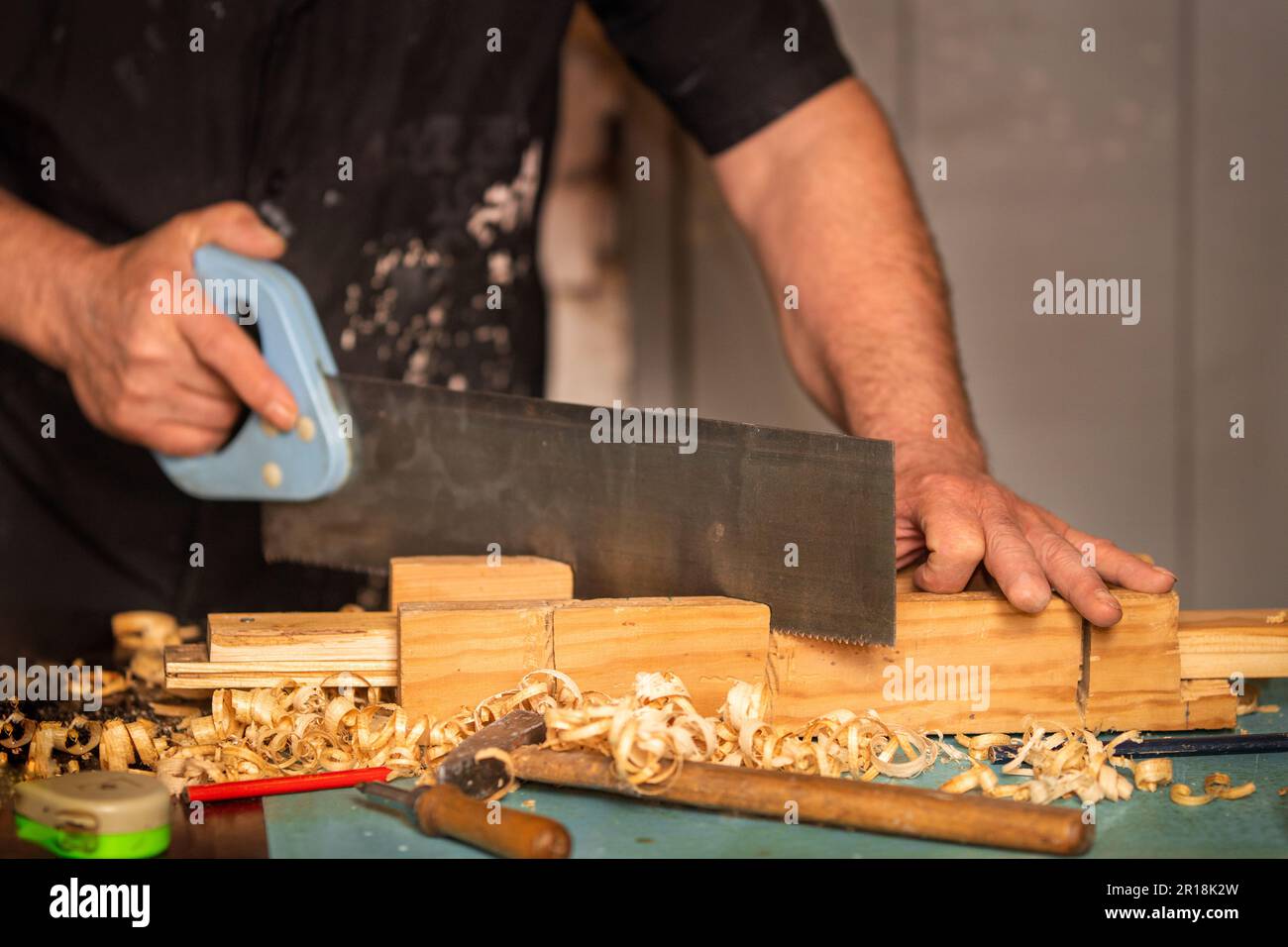 A carpenter saws wood with hand tools amidst a messy workspace. The ...