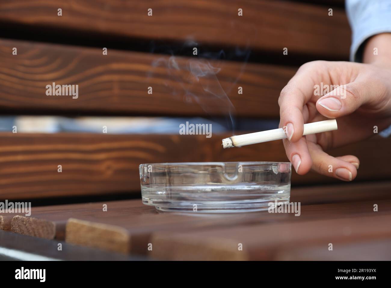Woman holding cigarette over glass ashtray on bench outdoors, closeup ...