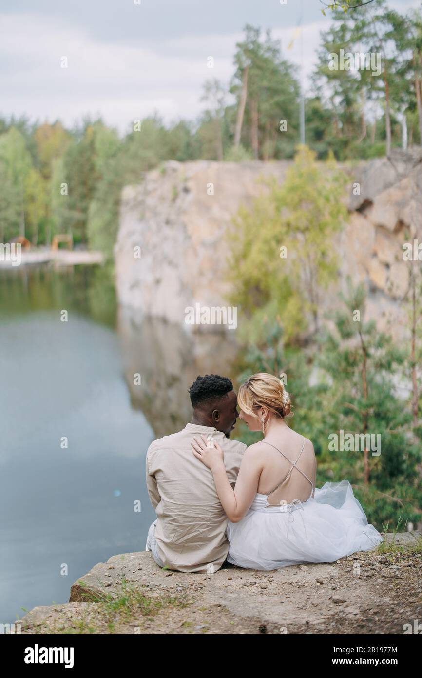 Happy interracial couple newlyweds sits on rock and embraces against ...