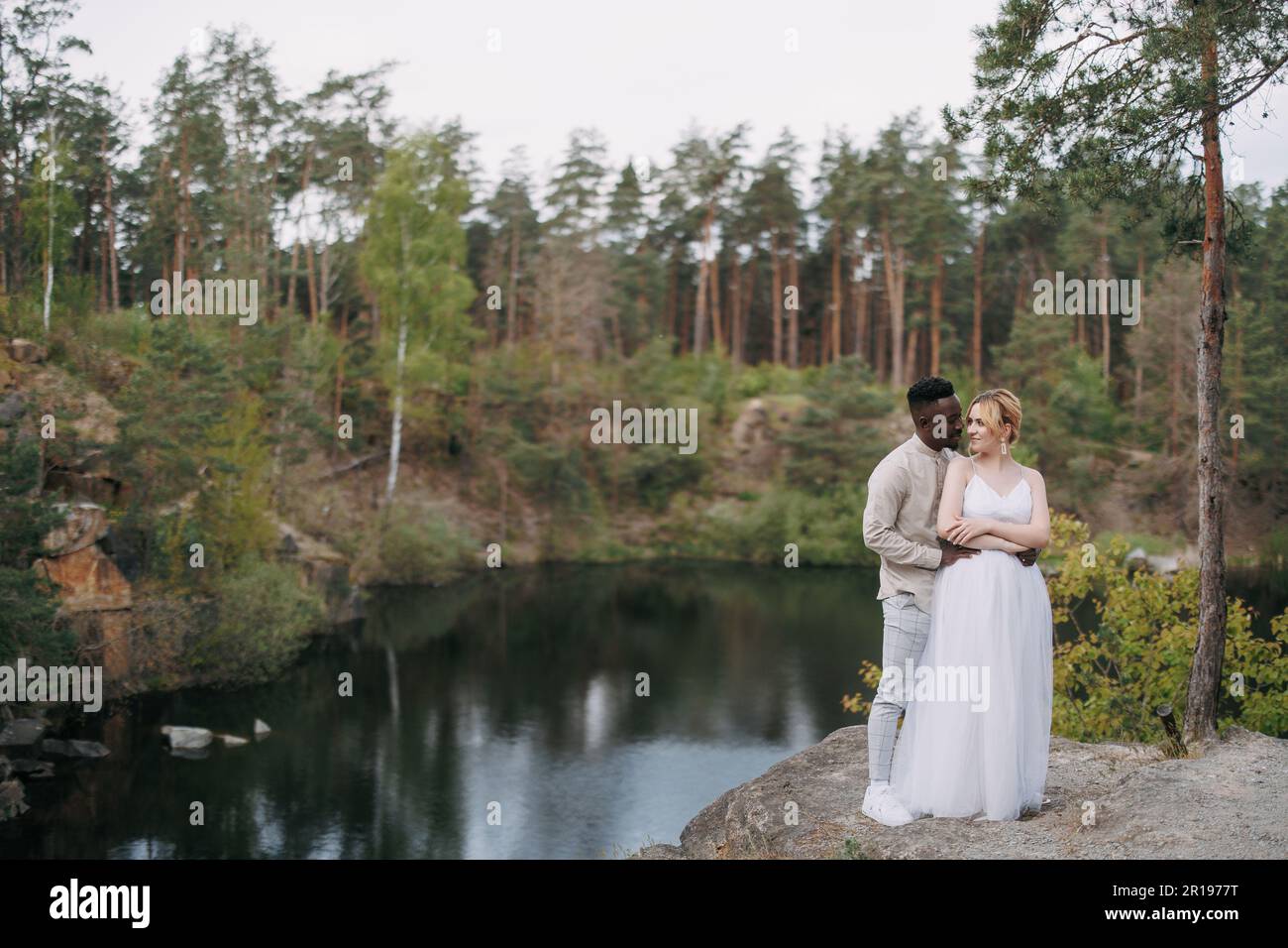 Happy interracial couple newlyweds stands on rock and embraces against ...