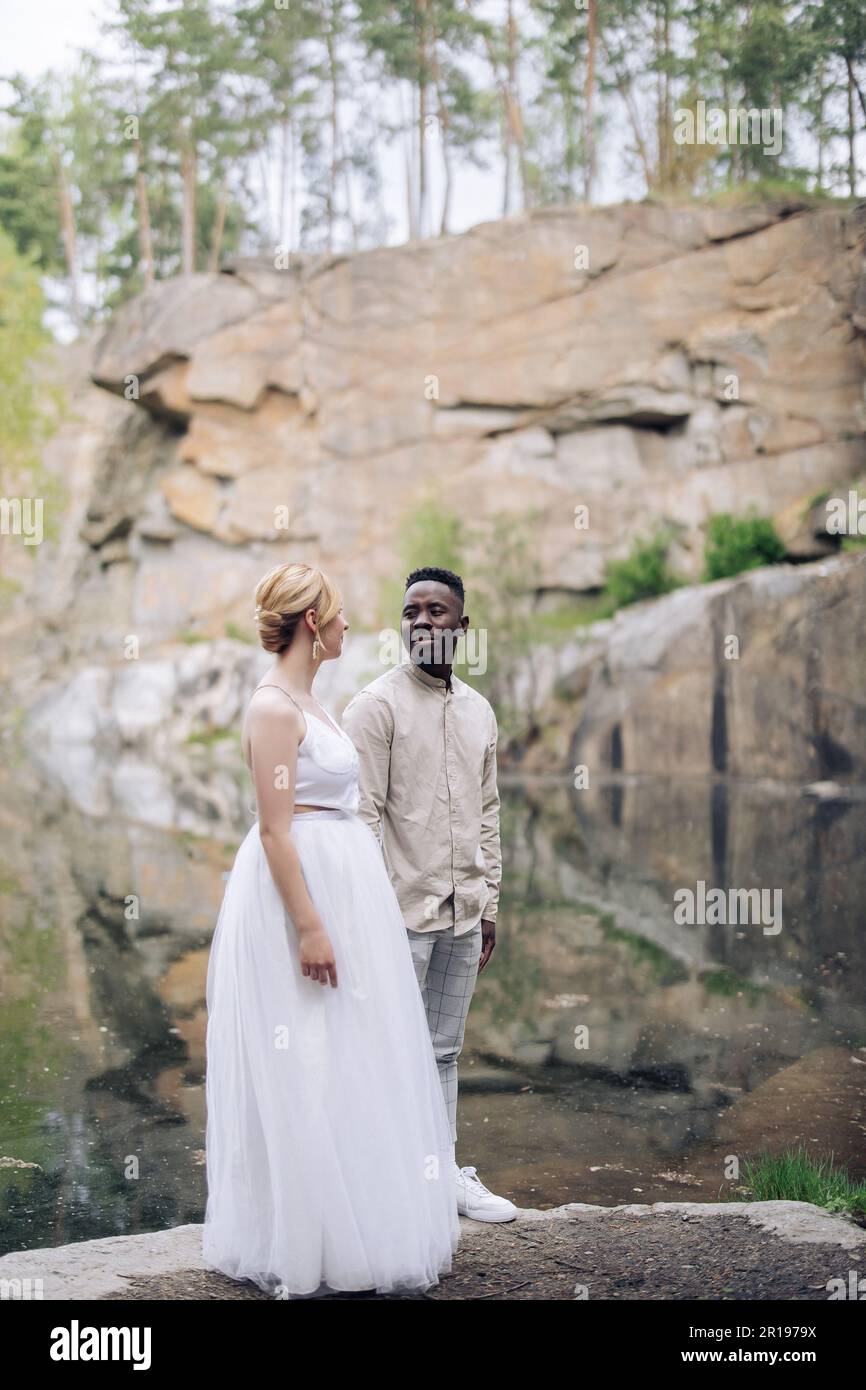 Happy interracial couple newlyweds stands on rock and looks each other ...