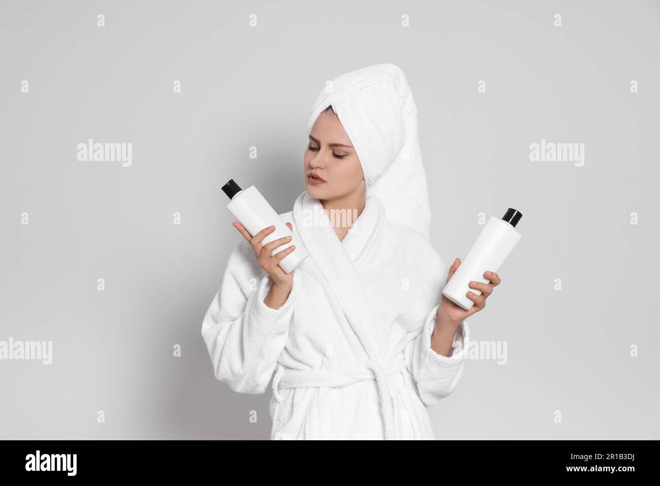 Beautiful young woman in bathrobe holding bottles of shampoo on light ...