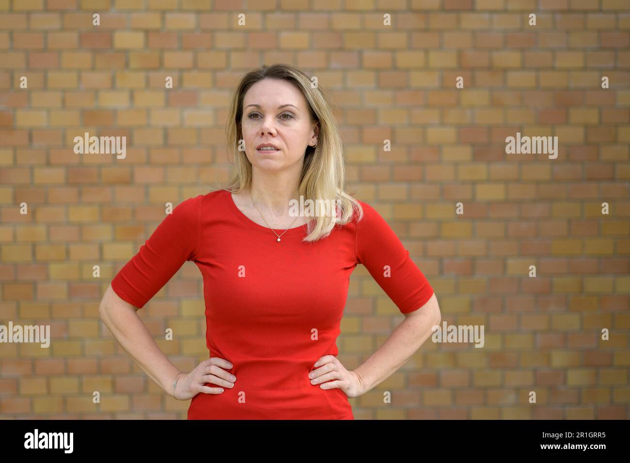 Pensive attractive middle aged blonde woman in front of a wall wearing a red top has her hands ...