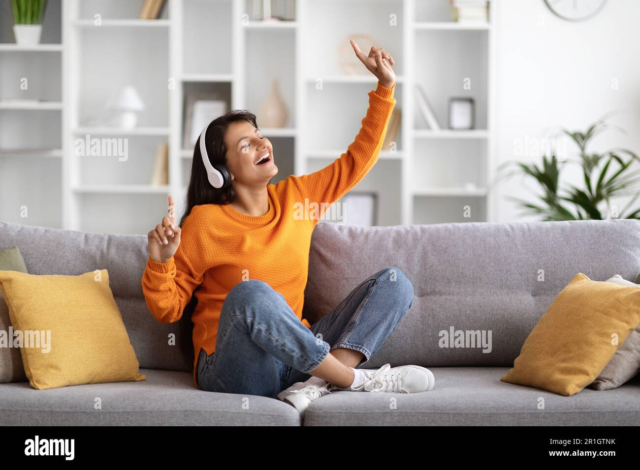 Joyful eastern lady sitting on couch, listening to music, singing Stock ...