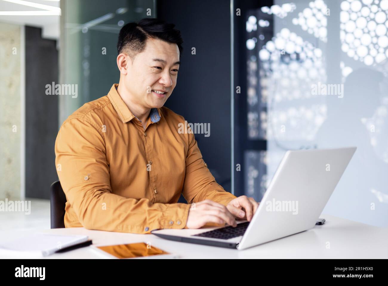 Cheerful and smiling Asian man working inside office, businessman in shirt and glasses using ...