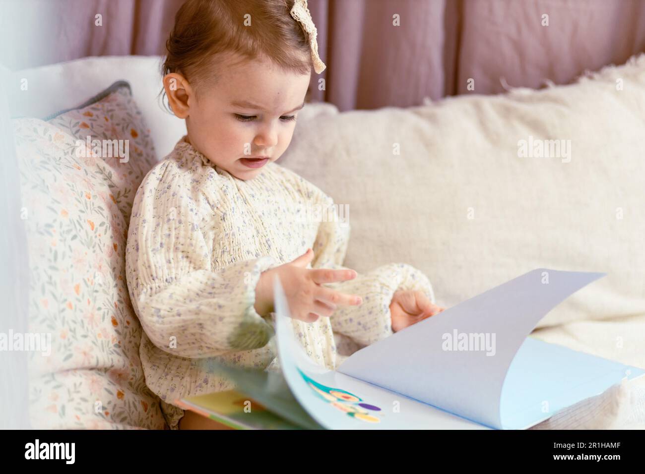 Cute, little baby girl sitting on the pillows reading a book, turning ...