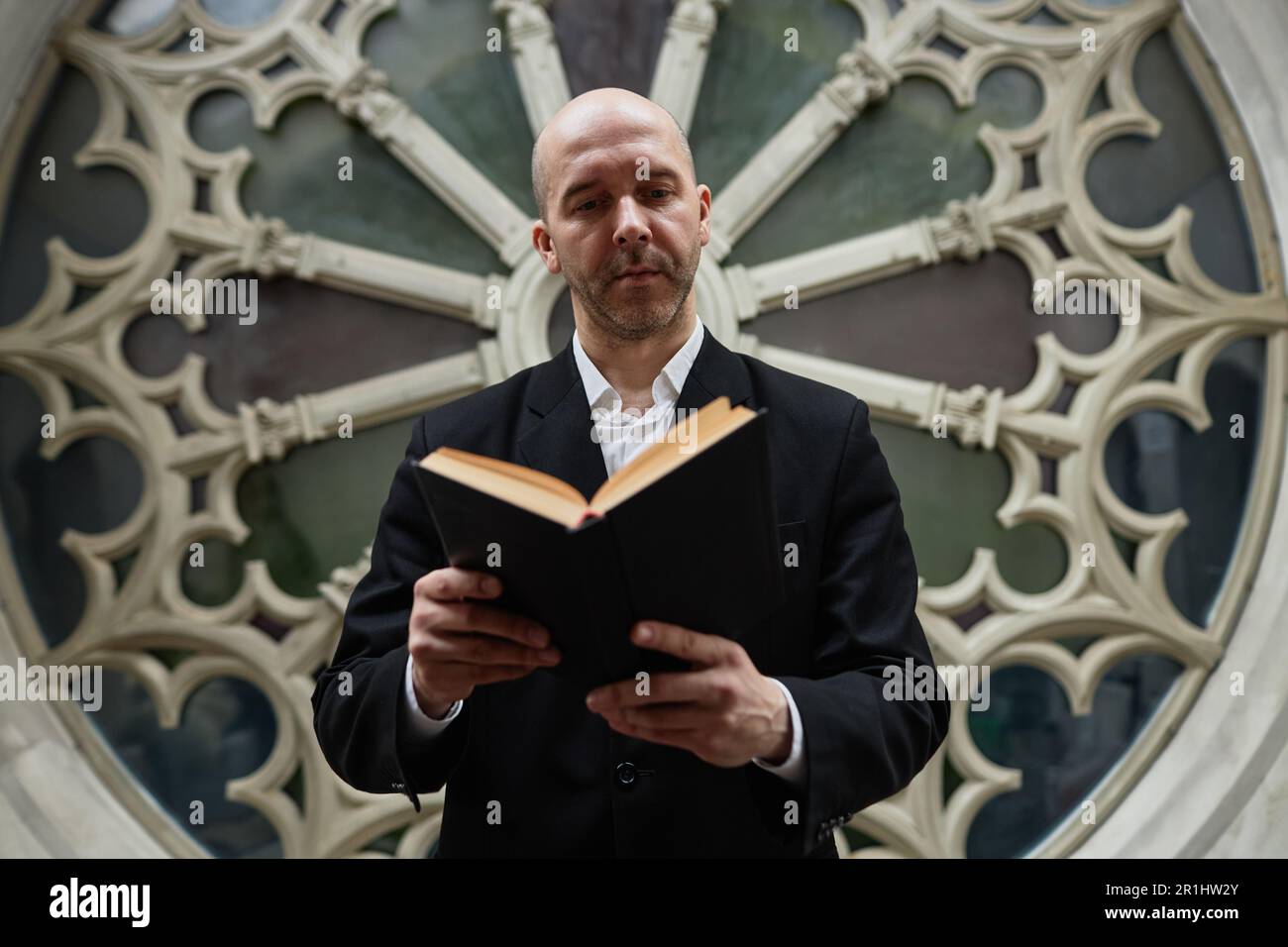 Mature pastor in black suit reading Bible while standing in church with ...