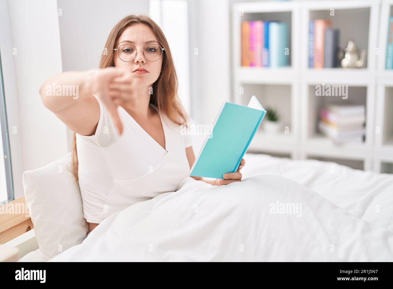 Young caucasian woman sitting on the bed at home reading a book with ...
