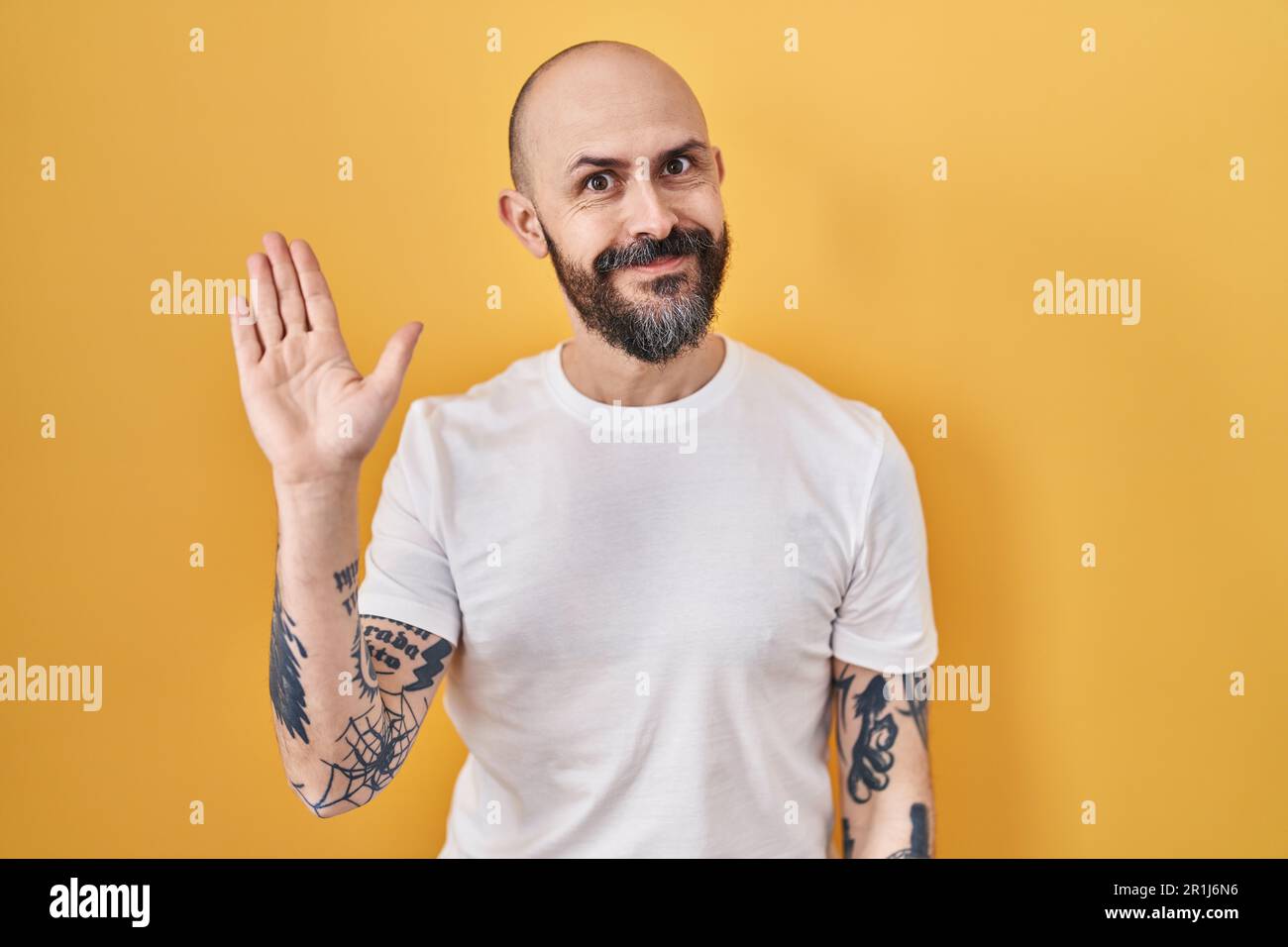 Young hispanic man with tattoos standing over yellow background waiving ...