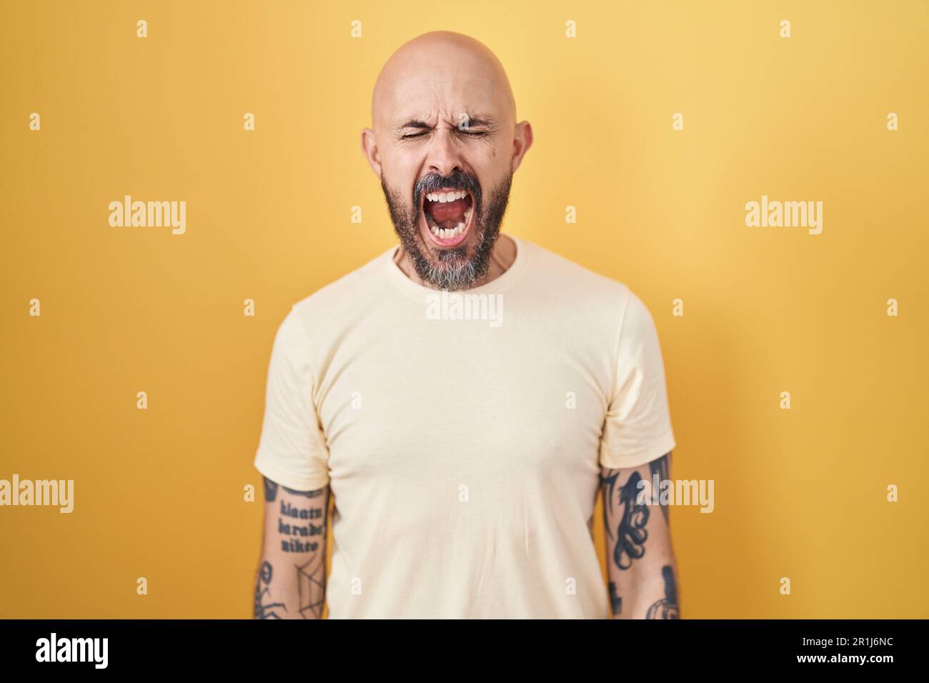 Hispanic man with tattoos standing over yellow background angry and mad ...