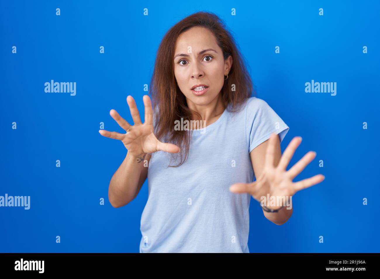 Brunette woman standing over blue background afraid and terrified with ...