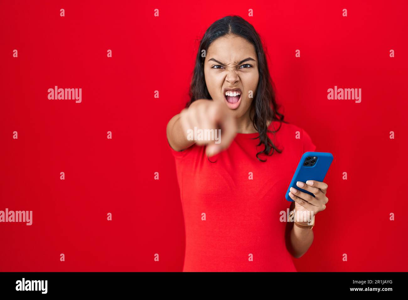 Young brazilian woman using smartphone over red background pointing ...