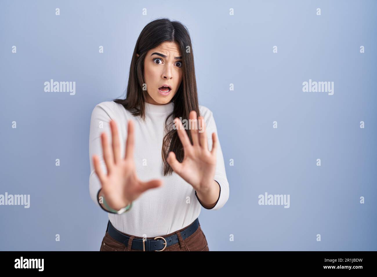 Young brunette woman standing over blue background afraid and terrified ...
