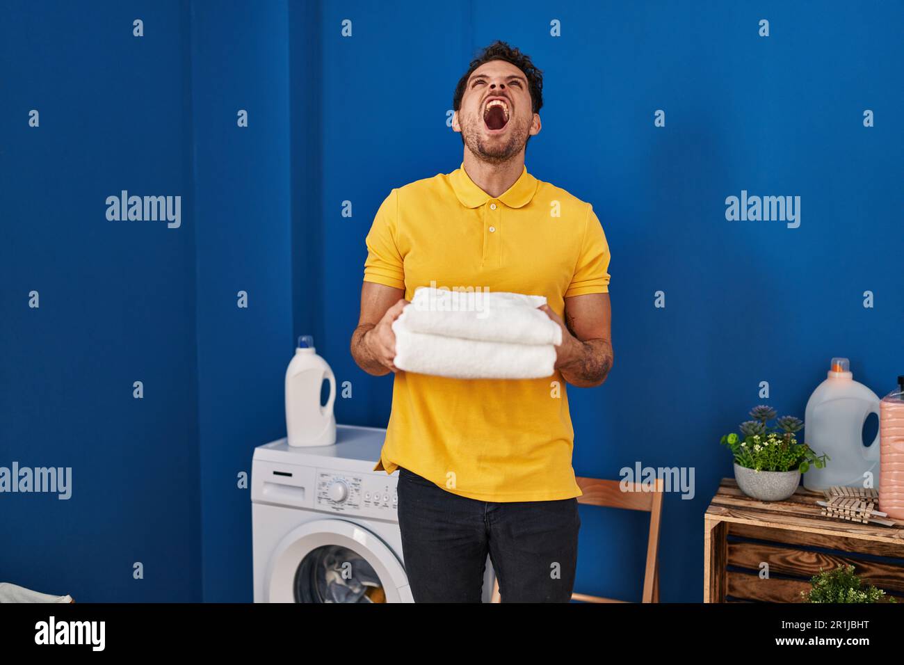 Young hispanic man holding clean towels at laundry room angry and mad screaming frustrated and ...