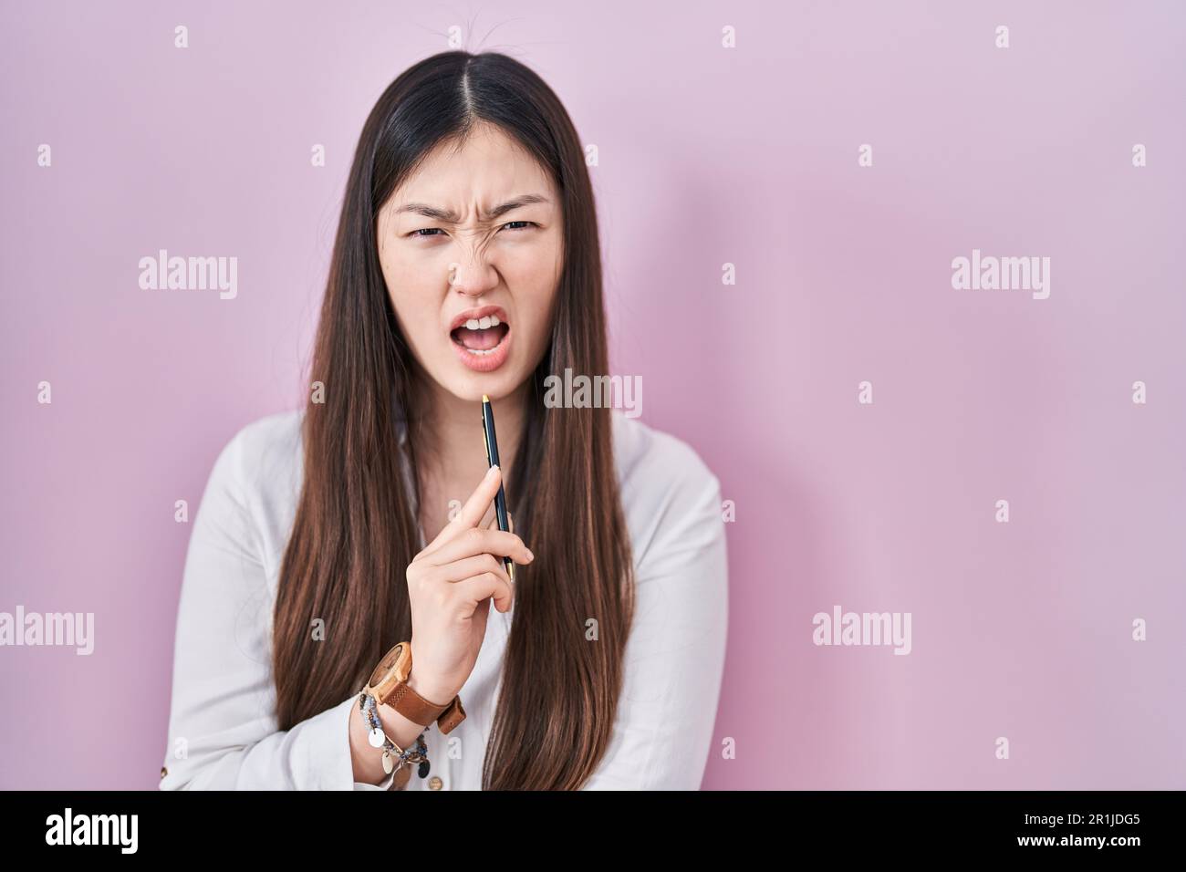 Chinese young woman holding pencil over pink background angry and mad ...