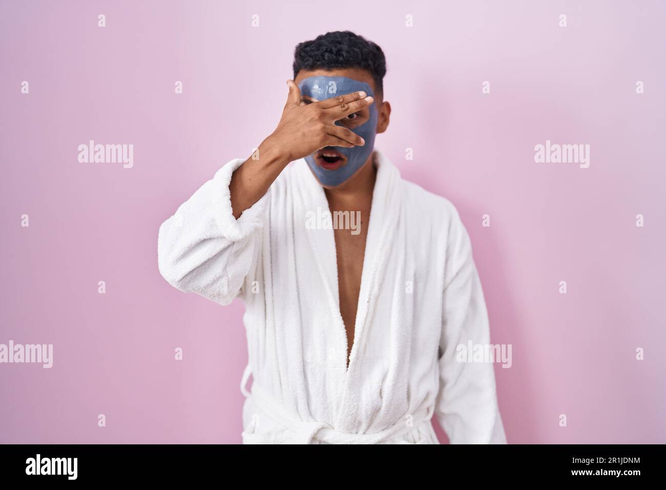 Young hispanic man wearing beauty face mask and bath robe peeking in ...