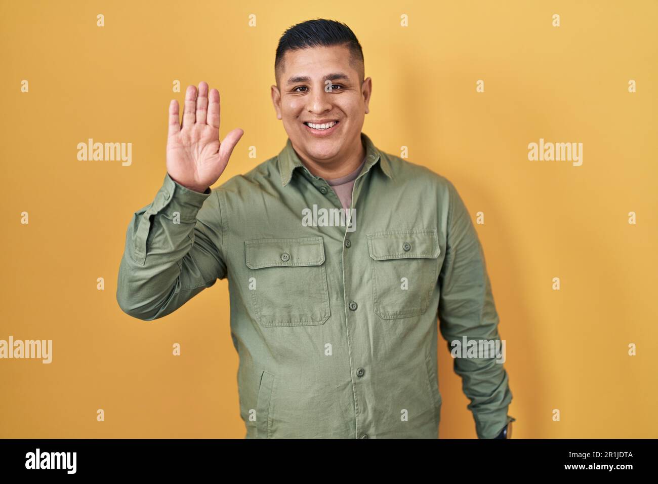 Hispanic young man standing over yellow background waiving saying hello ...
