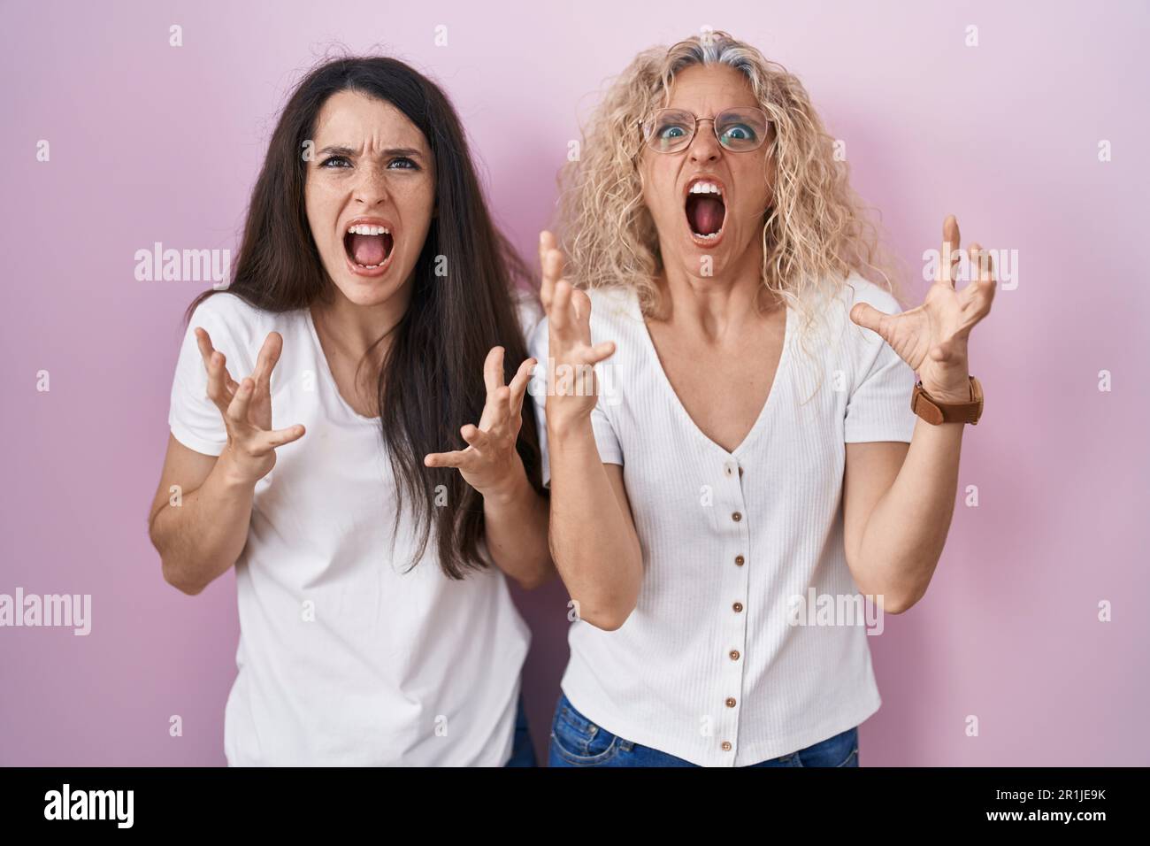 Mother and daughter standing together over pink background crazy and ...
