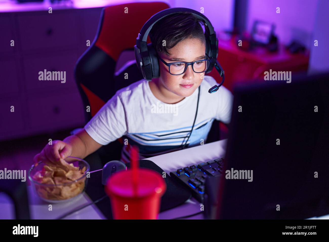 Adorable hispanic boy streamer playing video game eating chips potatoes ...