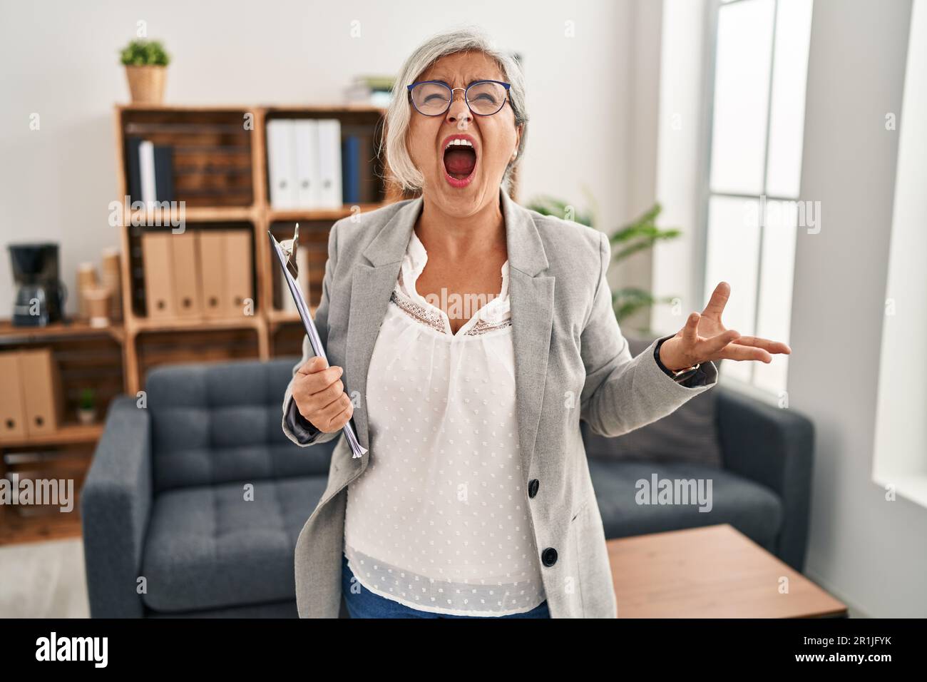Middle age woman with grey hair at consultation office angry and mad screaming frustrated and ...