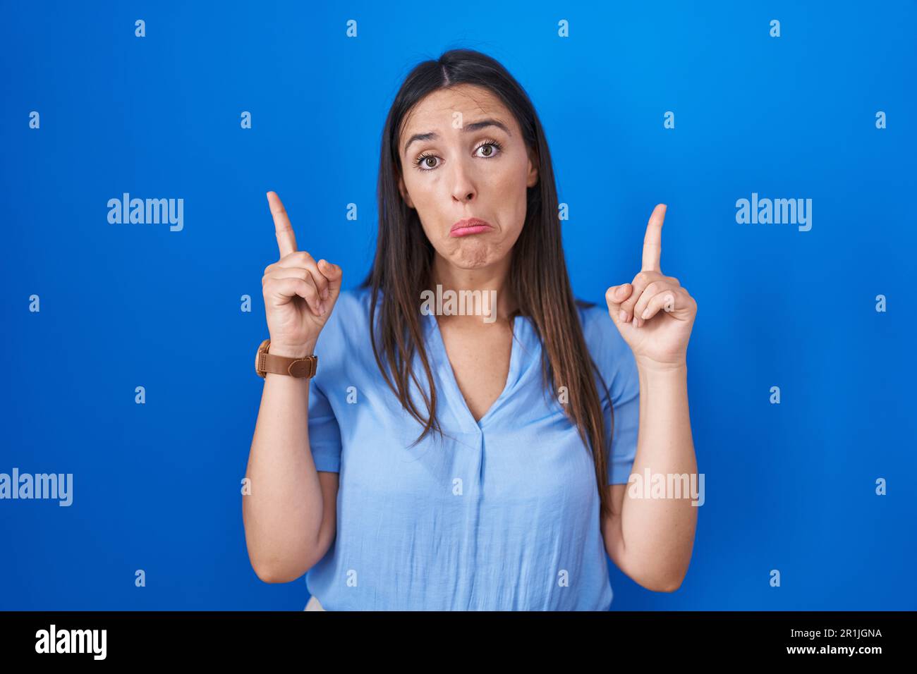 Young brunette woman standing over blue background pointing up looking ...