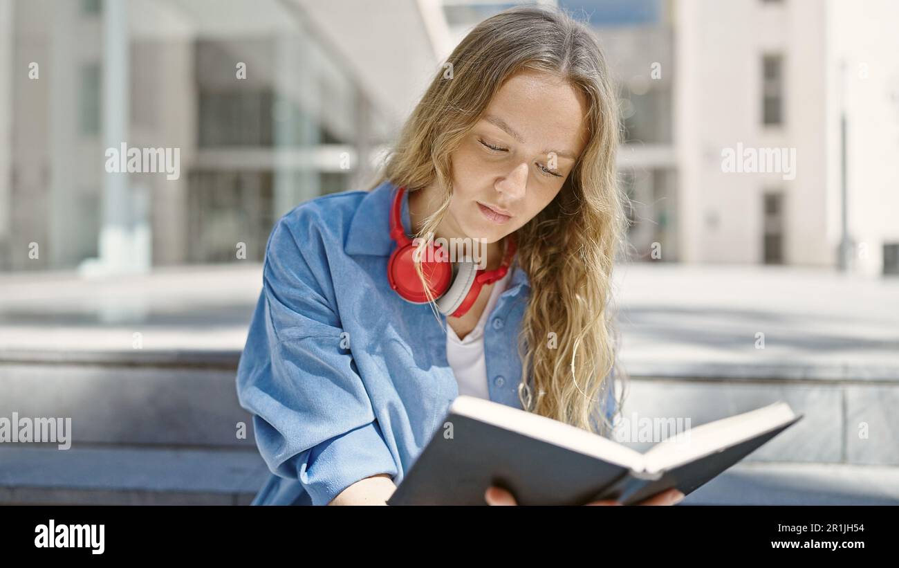 Young blonde woman student reading book sitting on stairs at university ...