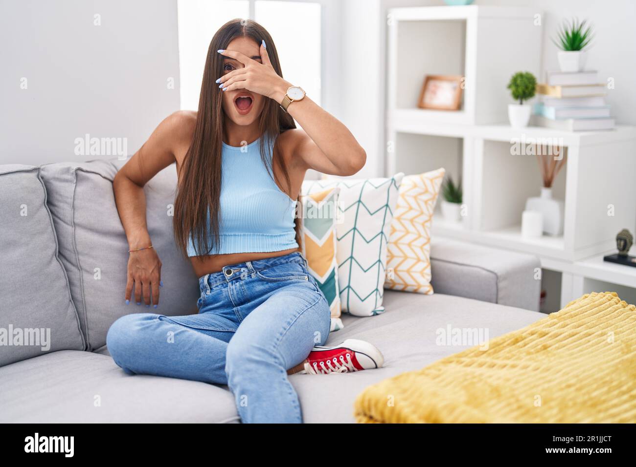 Young brunette woman sitting on the sofa at home peeking in shock ...