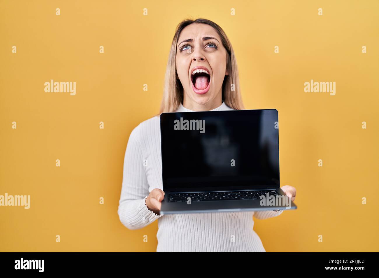 Young blonde woman holding laptop angry and mad screaming frustrated ...