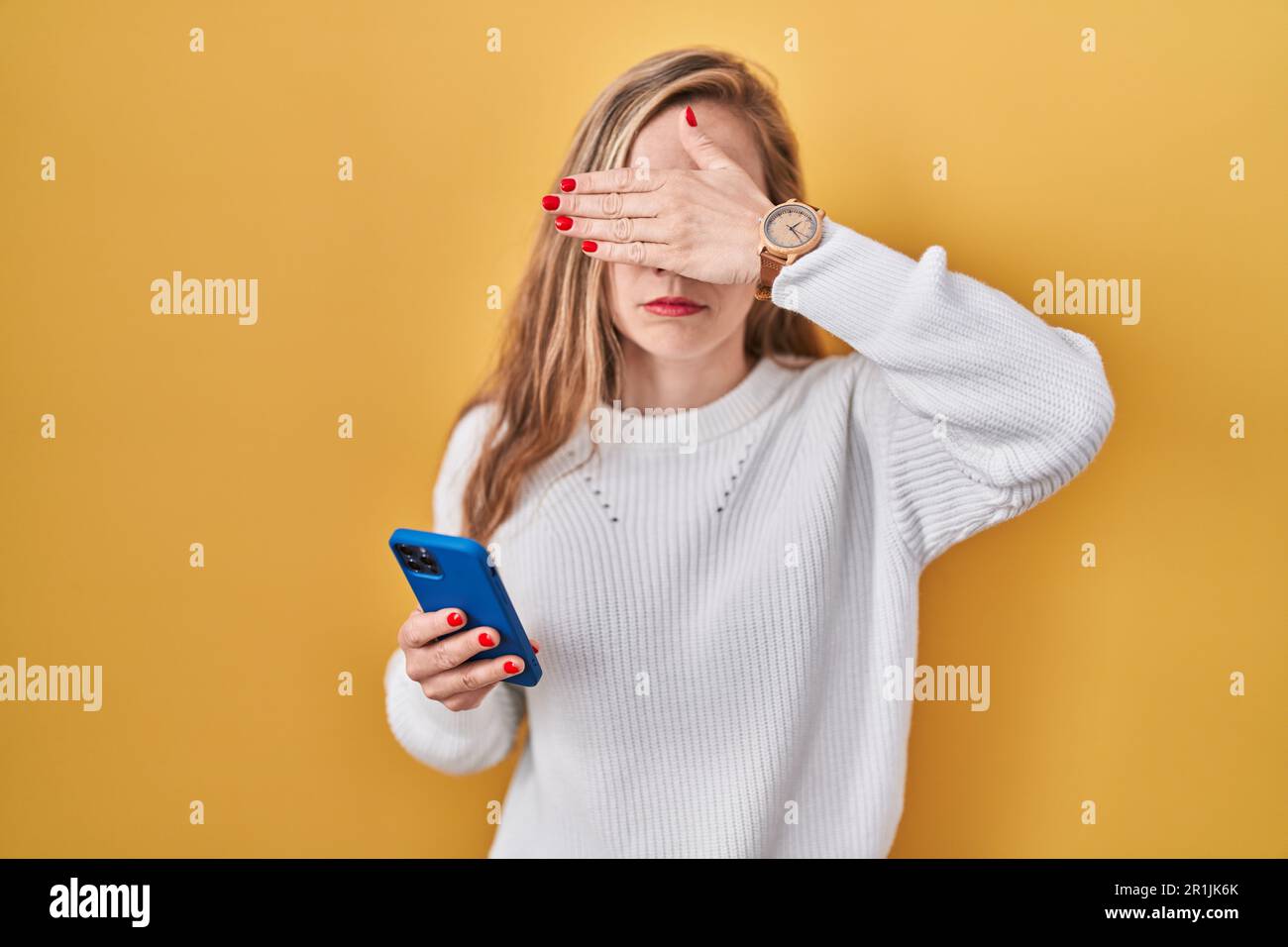 Young blonde woman using smartphone typing message covering eyes with ...