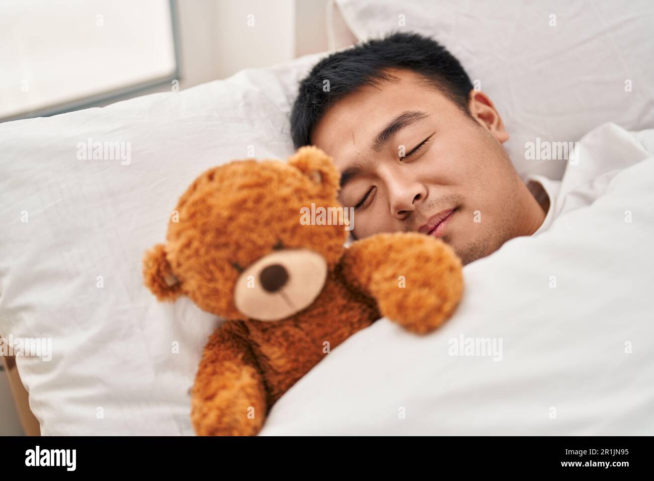 Young chinese man lying on bed sleeping with teddy bear at bedroom ...