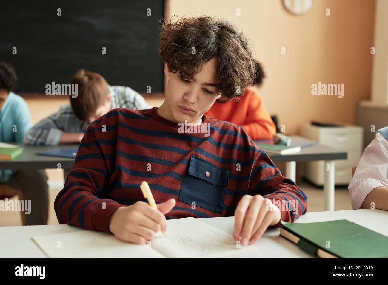 Portrait of teen boy with curly hair writing in notebook during school ...