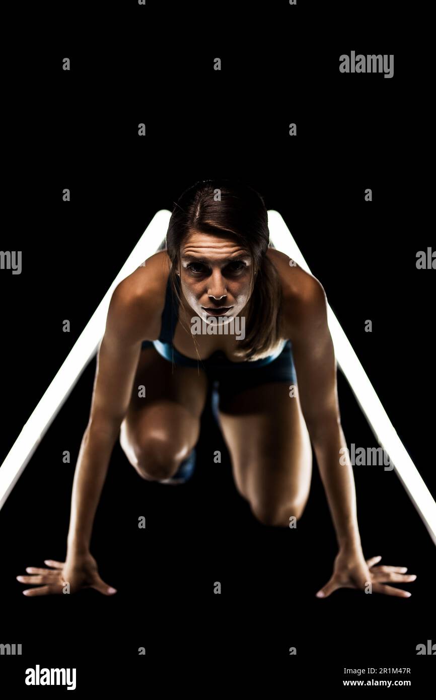 Female runner in race start position looking up. Girl in sportswear posing on lit track against ...