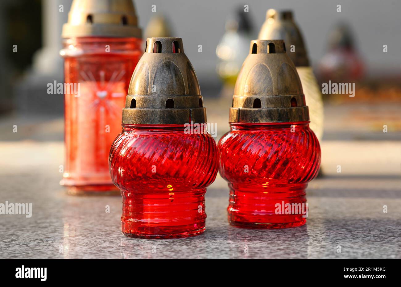 Different grave lanterns with burning candles on granite tombstone outdoors. Funeral ceremony ...