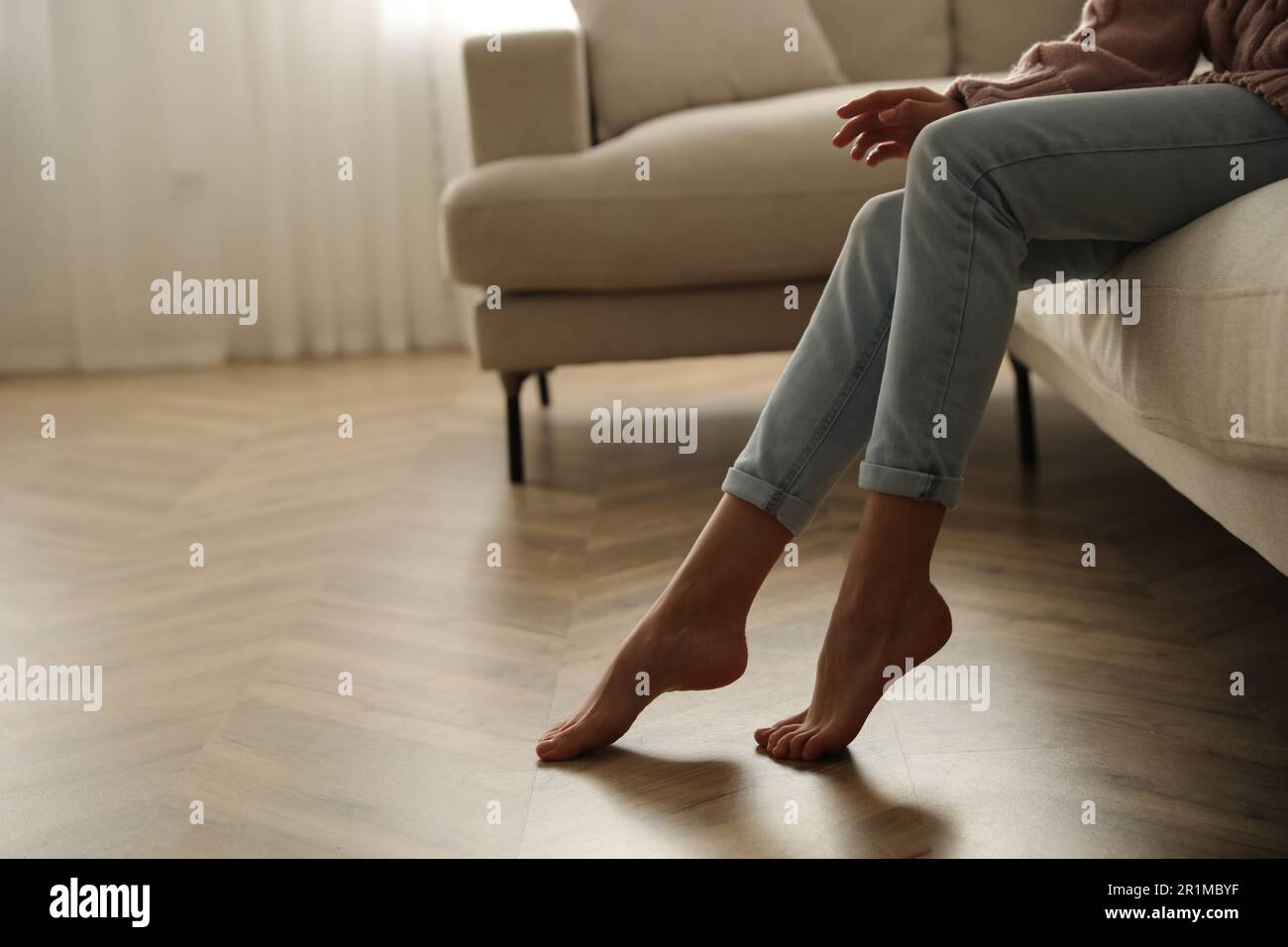 Barefoot woman sitting on sofa in living room, closeup. Floor heating ...