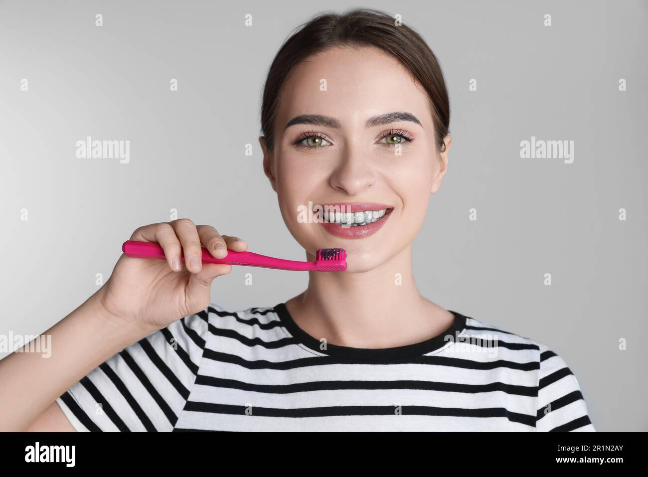 Young woman brushing teeth with charcoal toothpaste on grey background ...
