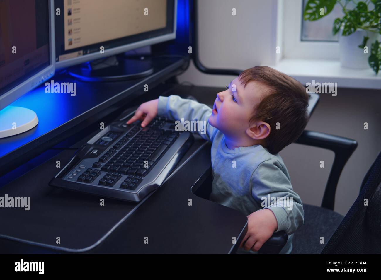 Happy baby is sitting at the computer keyboard next to the monitors. A ...