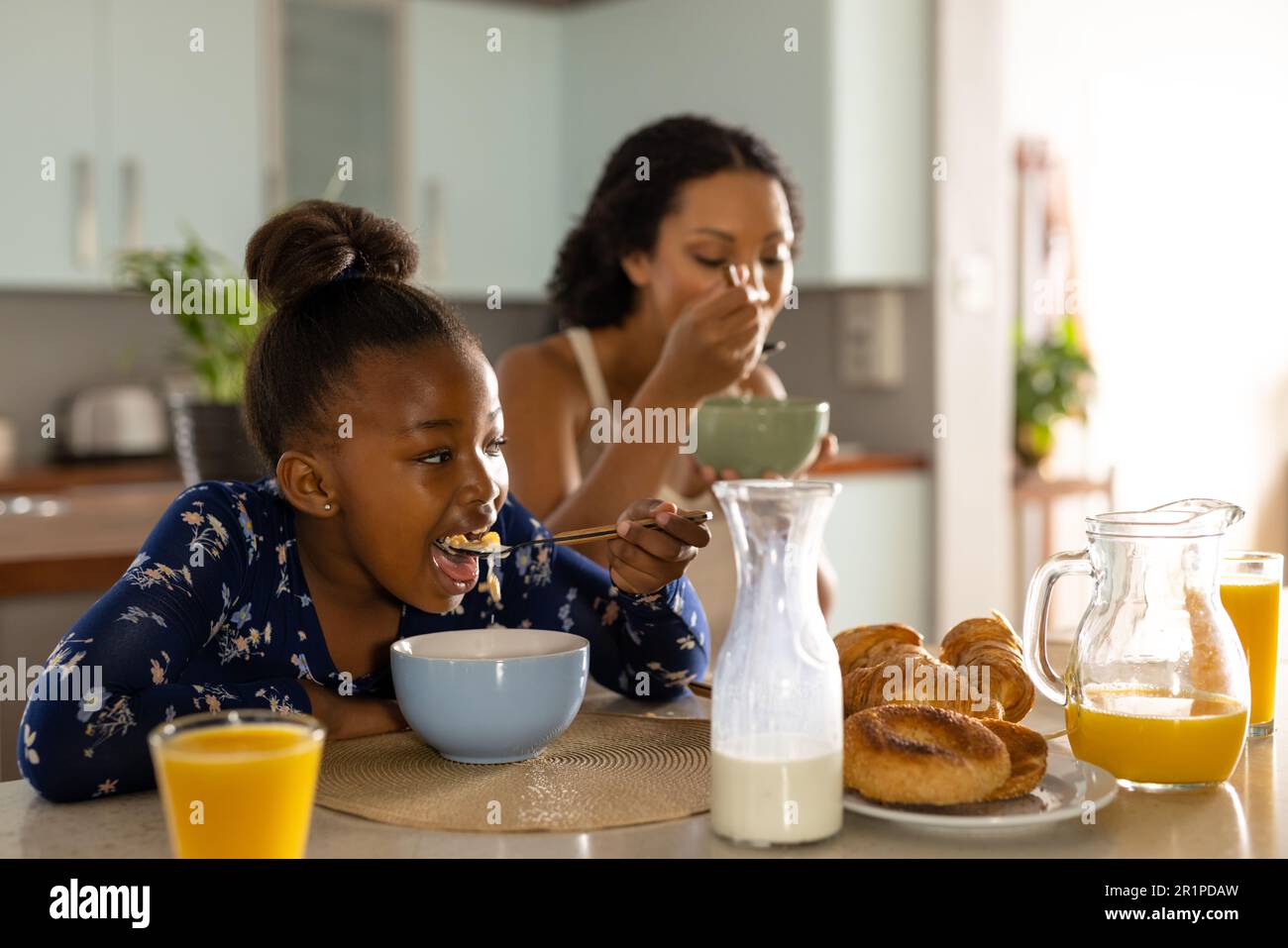 Happy african american mother and daughter sitting at kitchen island eating breakfast together ...