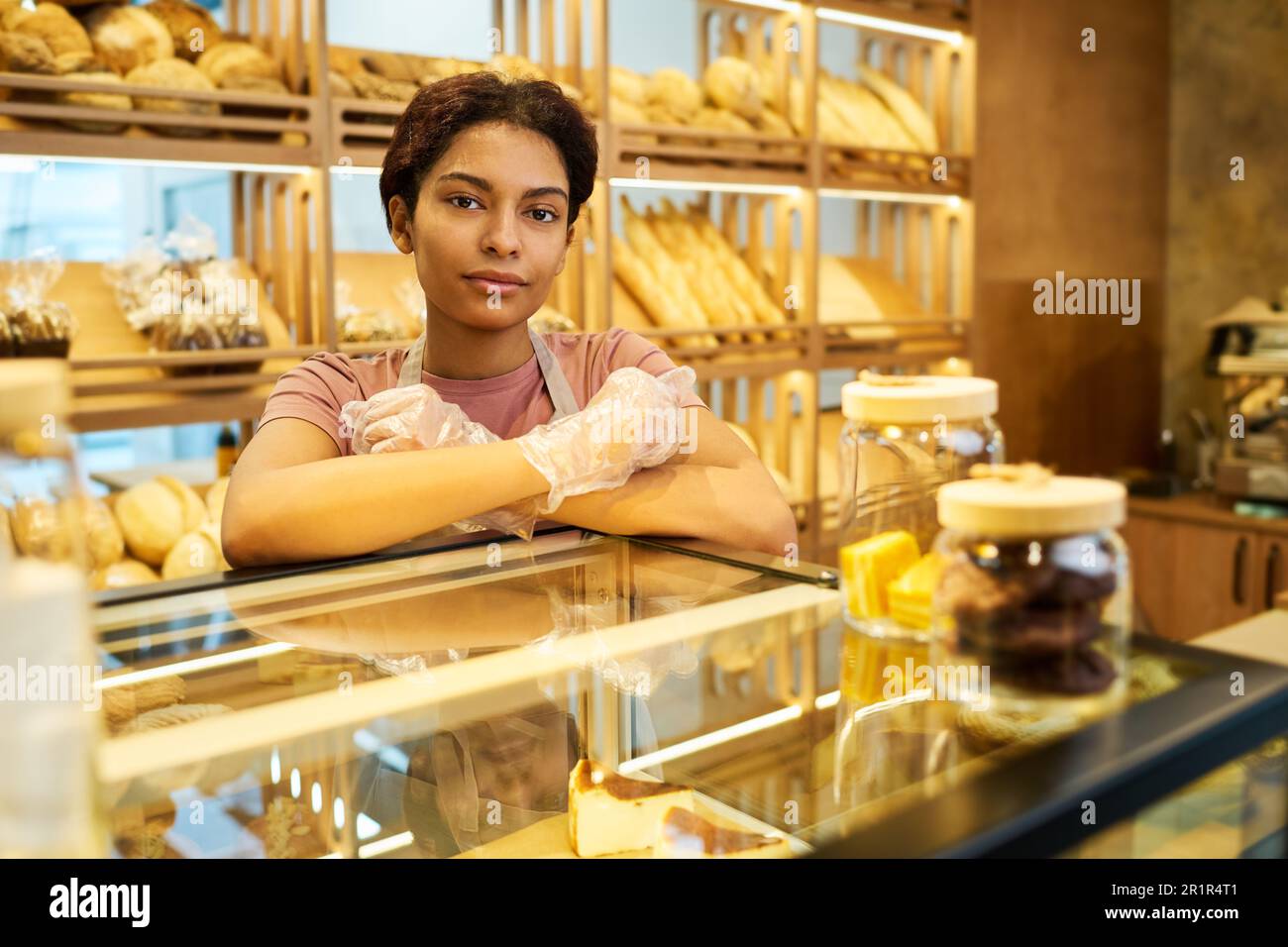 Young pretty female clerk standing by glass display with pastry in ...