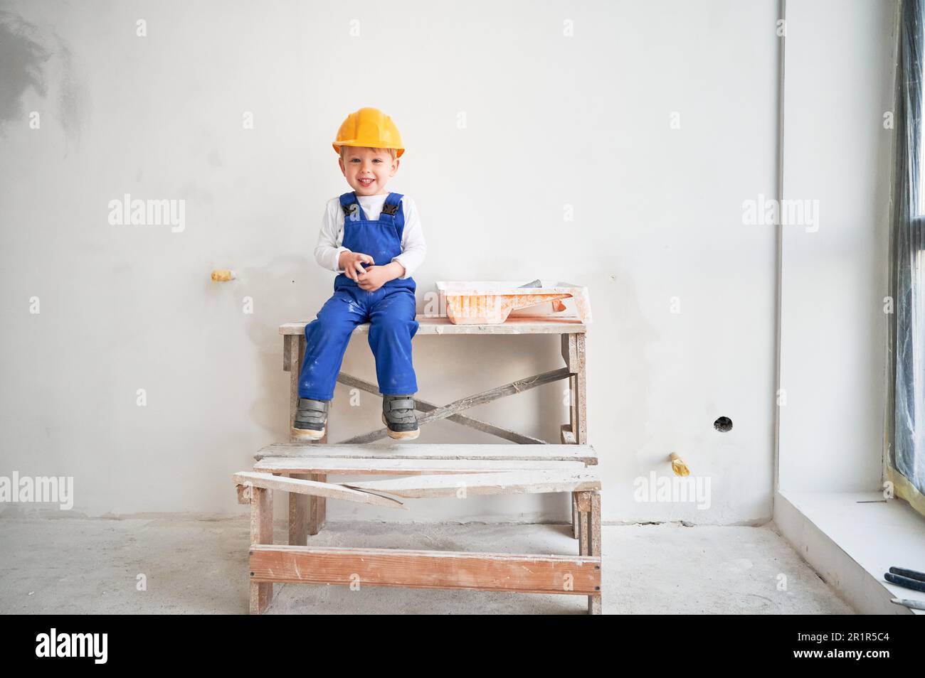 Cute kid construction worker sitting on wooden table against white wall ...