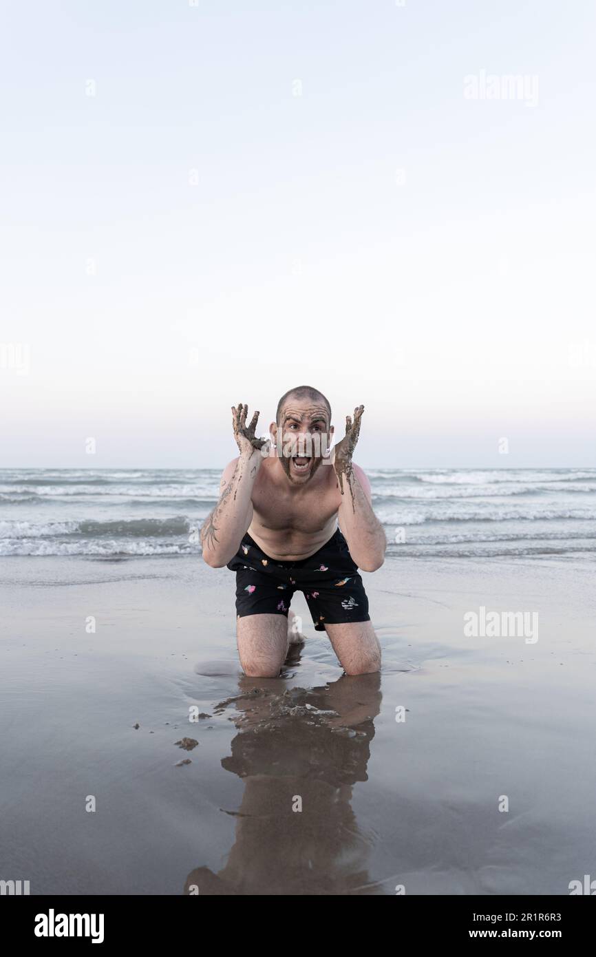 A young man is shouting desperately with beach sand in hands and face ...