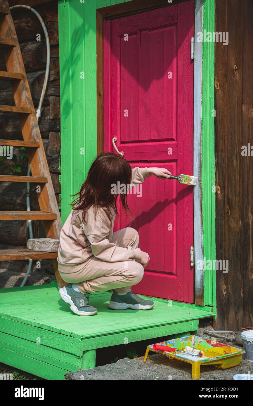 Cute girl painting the door of a wooden house in a bright purple, pink ...
