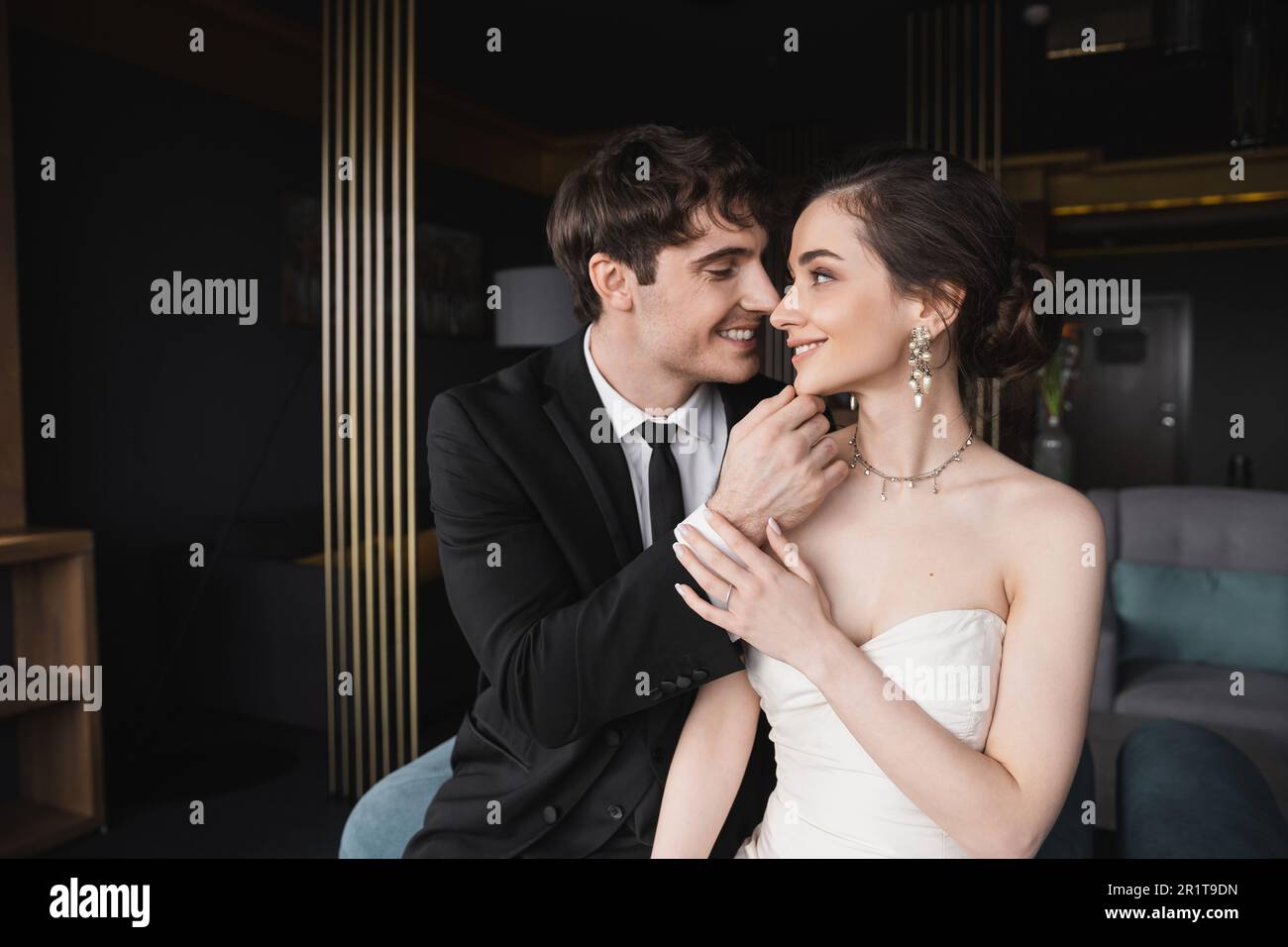 happy groom in black suit with tie touching face of charming bride in ...