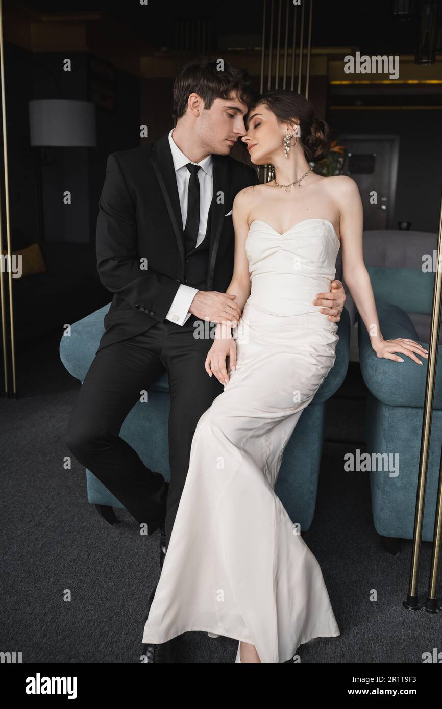 tender groom in black suit hugging waist of charming bride in white ...