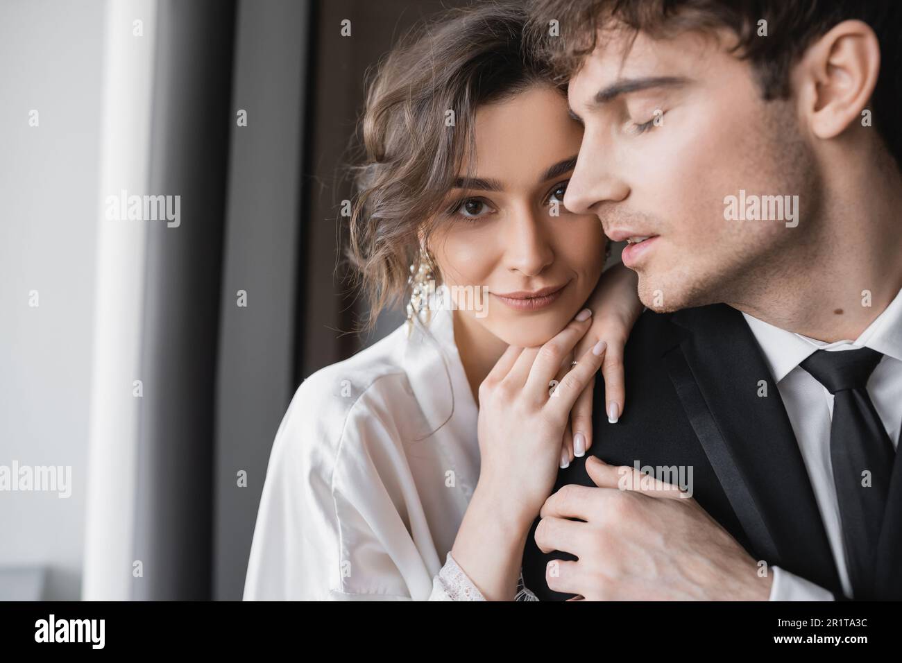 charming bride in jewelry and white silk robe leaning on shoulder of ...