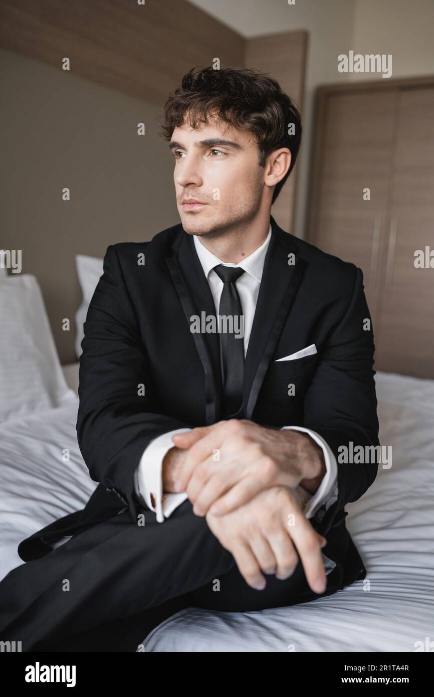 dreamy man in classy formal wear with black tie and white shirt sitting on bed in modern hotel ...