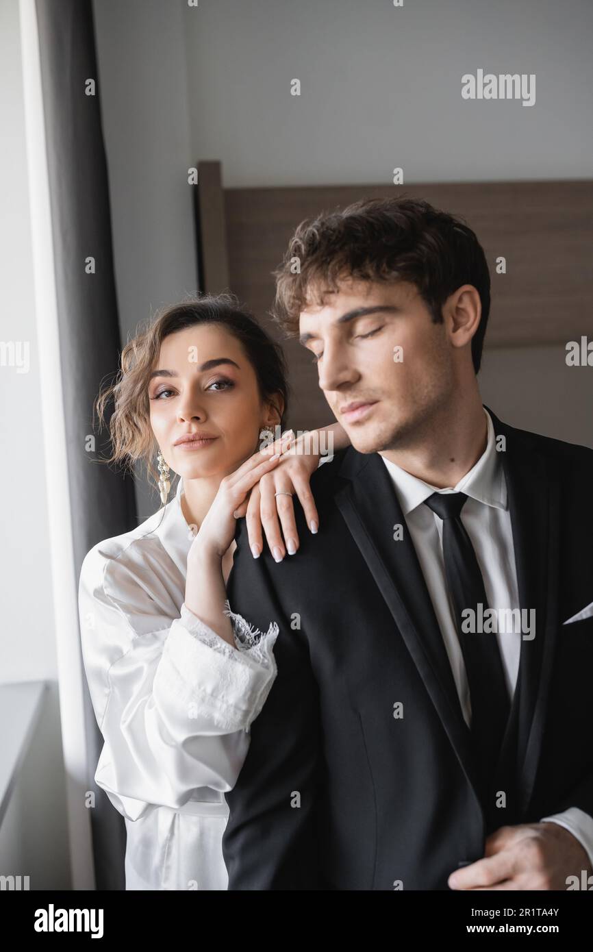 pretty bride in jewelry and white silk robe leaning on shoulder of ...