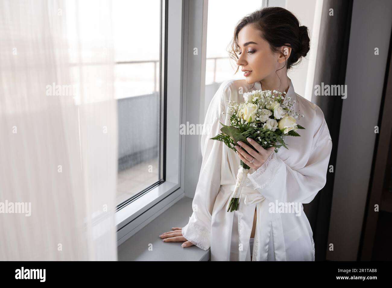 young woman with engagement ring on finger standing in white silk robe ...