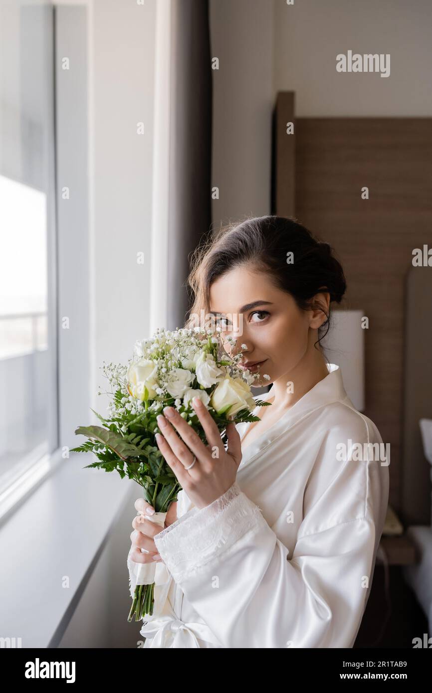young bride with engagement ring on finger standing in white silk robe ...