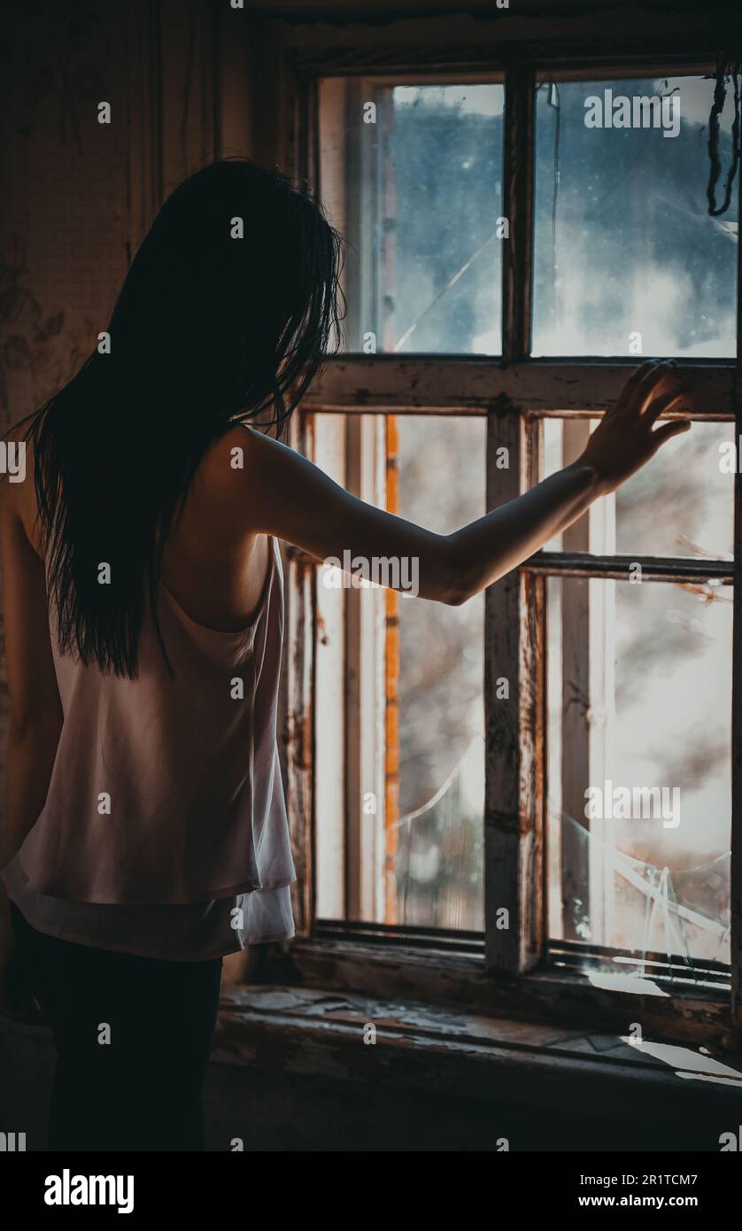 A lonely girl stands at a broken window and looks dreamily into the ...