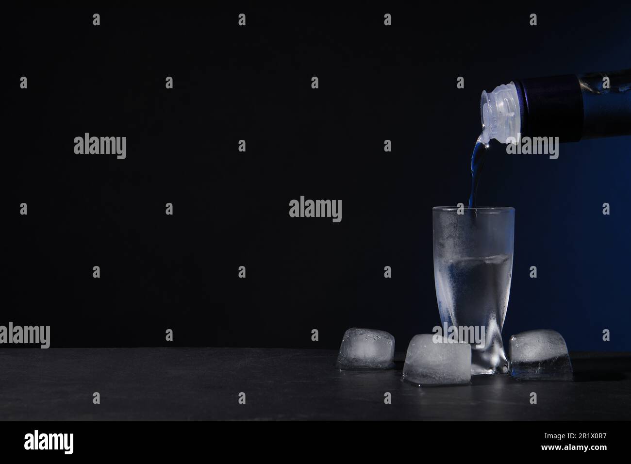Pouring vodka from bottle into shot glass on black table against dark ...